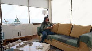 smiling latin woman sitting on the couch at home writing message on her cell phone, wellness at home