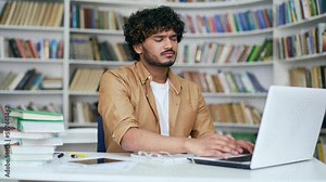 University student suffering from back pain while working on laptop while sitting in campus library space. Мale massages the lower back, rubs the muscles of the spine. He has sciatica or cold nerves