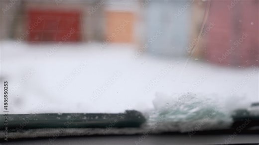 Close-up of a car's windshield wiper blade sweeping back and forth, cleaning away fresh snow and ice from the glass during a winter storm for better visibility while driving
