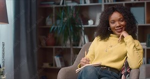Pretty african american female sitting in chair writing in notepad. Woman looking into camera smiling. Carefree people concept. Staying inside lockdown covid 19.