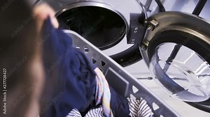 Interior of small laundromat in daylight. Close-up shot female holding basket , overhead view girl loading dirty clothes inside washing machine. Self-service. Public laundry