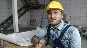 Medium shot of young Asian builder wearing hard hat and jeans overalls sitting in unfinished building and explaining how to draw layout plan before camera