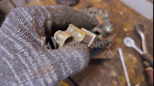 Professional auto mechanic wearing protective gloves holding and inspecting a vehicle's alternator voltage regulator unit, checking the brushes and overall condition in a repair shop