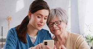 Smiling caregiver assists elderly patient with using a mobile phone, internet, and online technology, explaining and demonstrating app usage. Communicating and smiling, providing patient assistance.