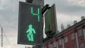 Close view of a digital traffic light showing a countdown with a green pedestrian signal, with a red city building and a cloudy sky