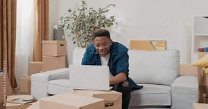 Young smiling boy sitting on the sofa in the new apartment after moving, checking emails on the laptop, good news, joy, finalizing the order, happiness hands up victory