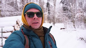 Man traveler with glasses unbuttons his jacket in the winter forest. He pauses on foot, recuperating. The snow lies on the plants in the bokeh. Camera movement.