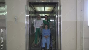 Two Caucasian young nurses are transporting old female patient in wheelchair, they are coming out of elevator and walking through the hospital building hallway, steadicam tracking shot