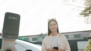 Slow motion young woman check battery status on smartphone while recharging EV car battery from charging station at city mall parking lot. Modern woman go shopping by electric vehicle. Expedient