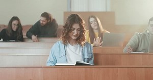 Young pretty female student reading textbook in classroom at seminar and talking with male groupmate by whispering. Educational concept. Girl and guy having chat at lection in college while read book.