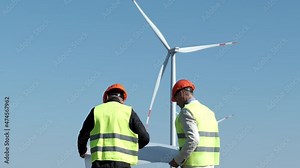 Wind turbines generate alternative energy at offshore station. Professional workers look at project drawing against rotating propellers backside view