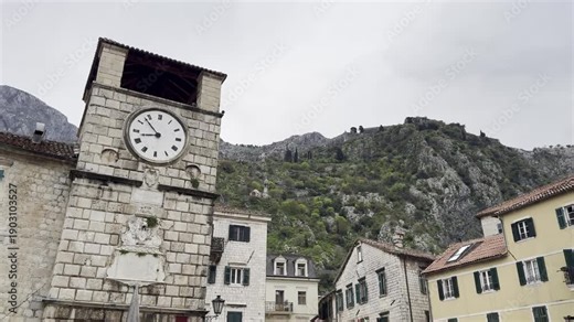 View of the historic clock tower in Kotor Old Town with stone buildings and the walls of Kotor Fortress rising on the hillside above - Kotor, Montenegro