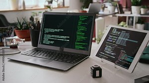 No people closeup of desk of programmer in office of IT company - laptop, tablet with running computer code being performed on black screen, potted plants and personal belongings