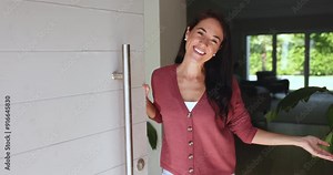 Cheerful young woman opens door, standing at doorway of modern apartment, warmly greets friend or neighbour with smile, extended hand showing welcoming gesture, invites invitee inside her cozy flat