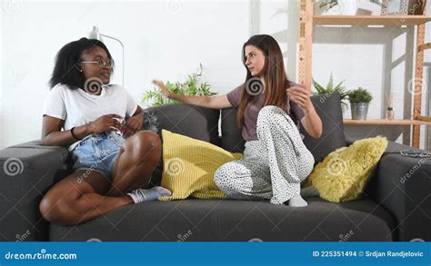 Two Young Female Roommates From College Are Sitting At Home On The Sofa And Talking About The