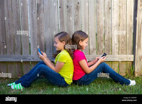 Twin Sister Girls Playing With Smartphone Sitting On Backyard Lawn Fence Leaning On Her Back