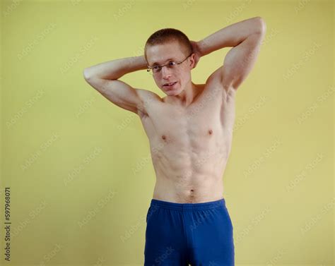 Young Naked Man In Glasses And Blue Shorts Holds His Hands Behind His Head On An Isolated Yellow